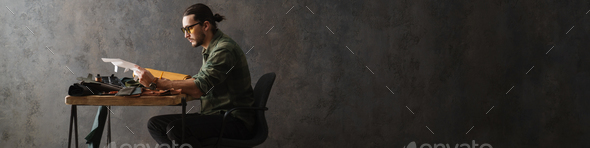 Bearded focused craftsman writing down notes while working with leather ...