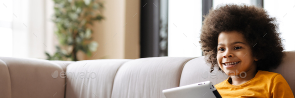Black curly boy smiling and using tablet computer while sitting on sofa ...