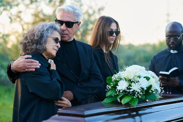 Mourning family of three and priest in black attire standing in front ...