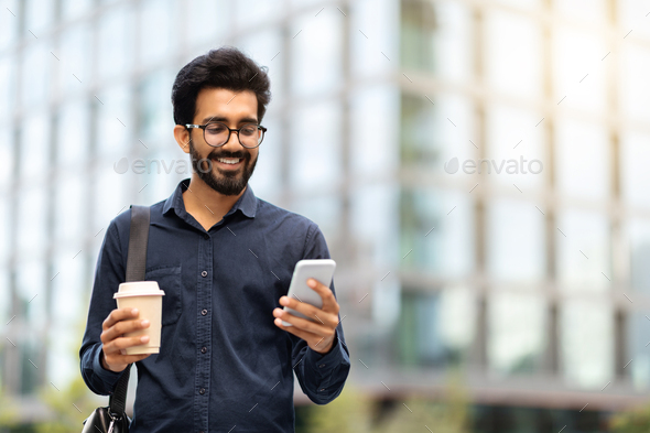 Cheerful indian guy going to office, using phone, drinking coffee Stock Photo by Prostock-studio