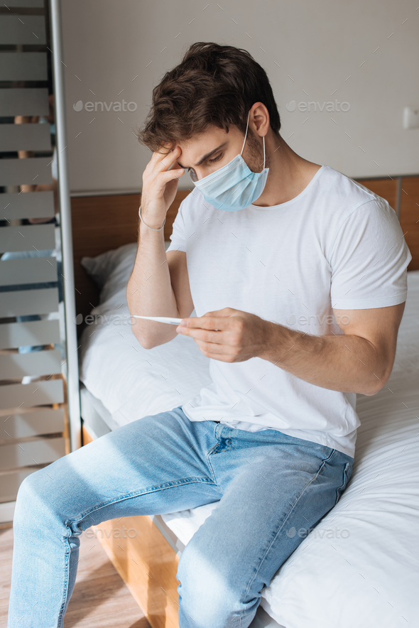tired ill man in medical mask looking at thermometer during self ...
