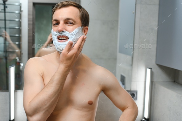 Young smiling Caucasian shirtless man applying shaving foam on face in front of mirror Stock ...