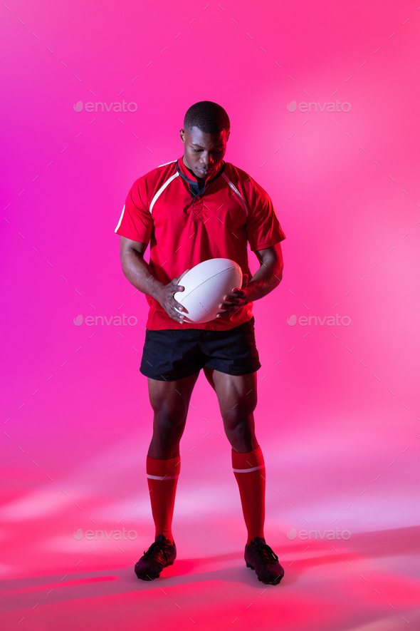 African american male rugby player with rugby ball over pink lighting ...