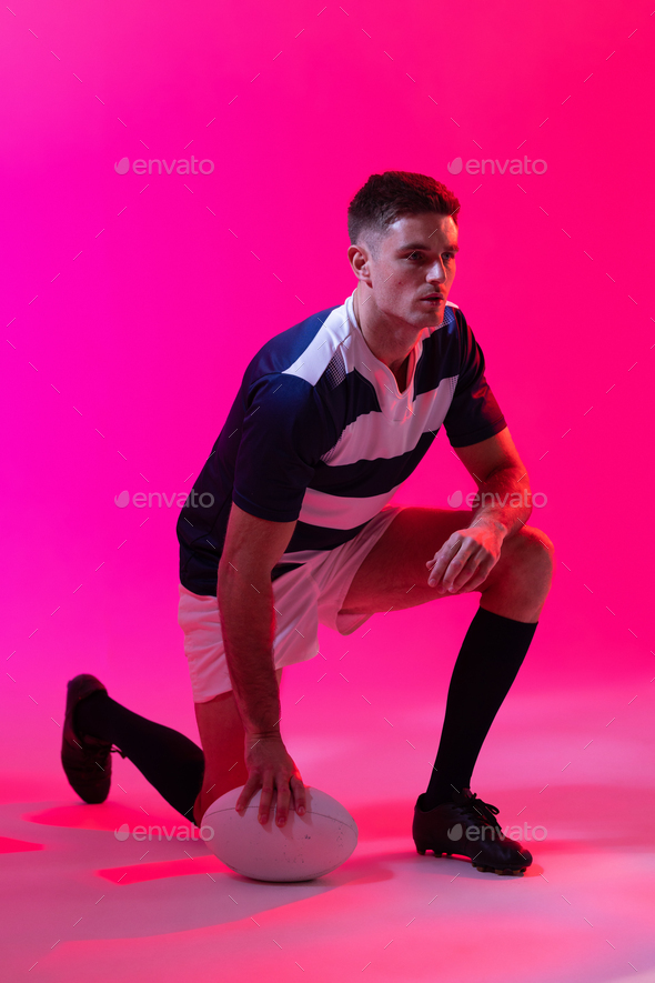 Caucasian male rugby player crouching with rugby ball over pink ...