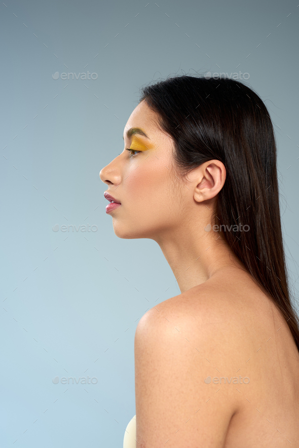 side view of model with bold makeup and bare shoulders posing on blue ...