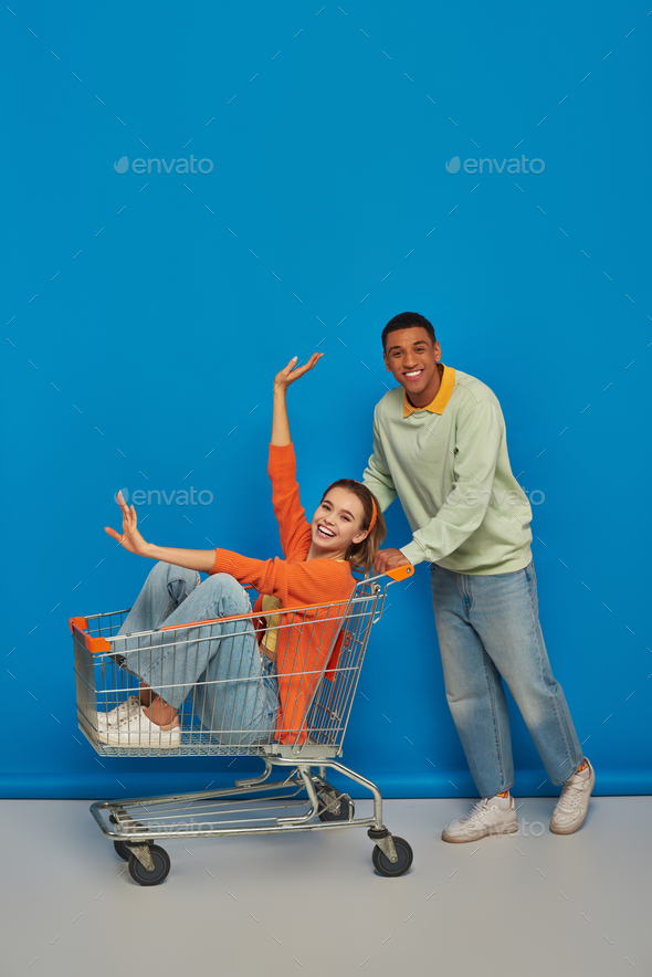 joyful african american man riding shopping cart with happy girlfriend ...