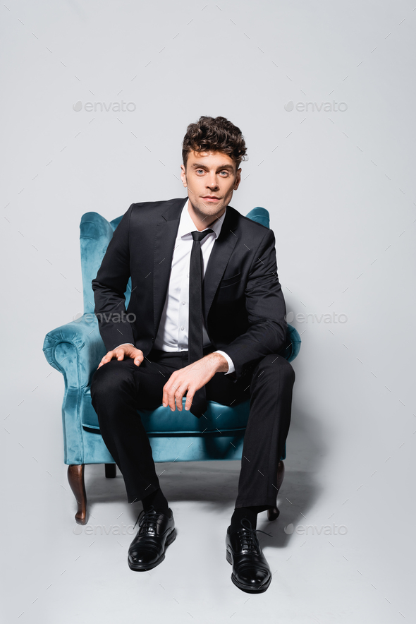 elegant man in suit sitting in velour armchair isolated on grey Stock ...