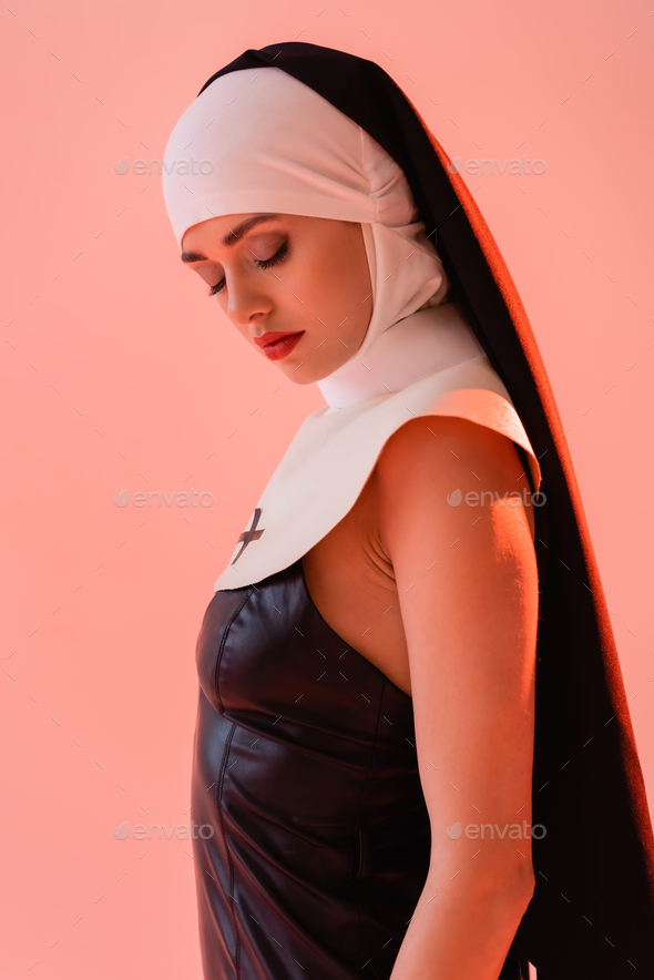 sensual nun in leather dress standing with closed eyes isolated on pink ...