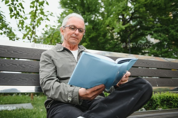 Old man with gray hair reads a book on a bench in the park. Rest in the ...
