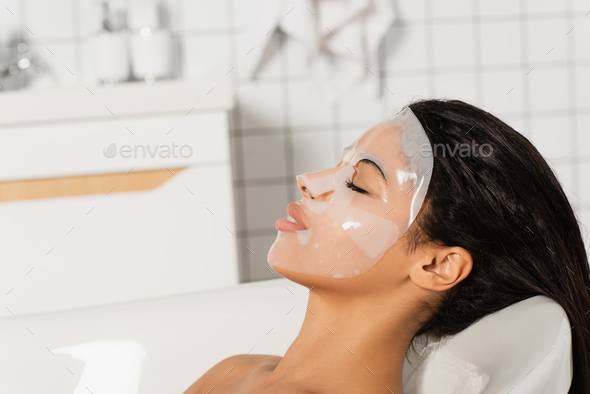 young african american woman with sheet mask on face lying in bathtub ...