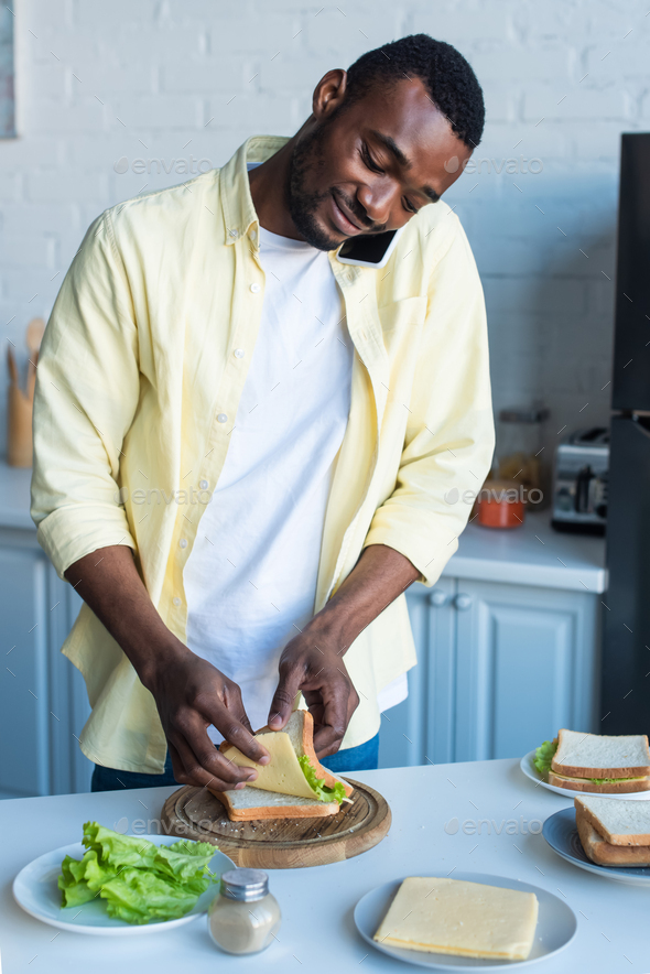 smiling african american man making sandwiches and talking on ...