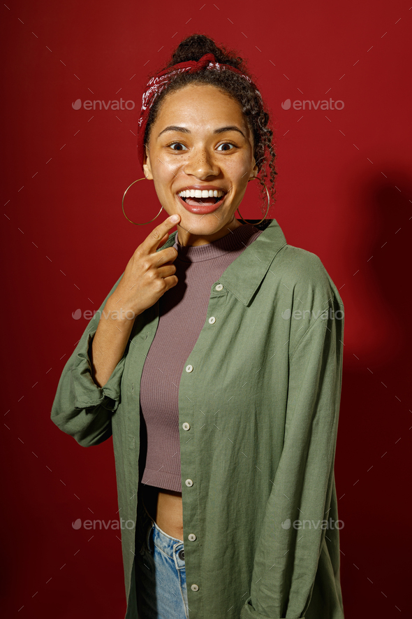 Pretty woman is touching cheek and looking camera with smile on red ...