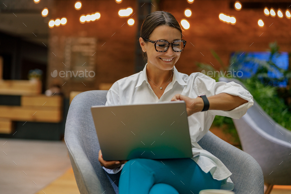 Female manager looking time on wrist watch while working laptop on ...
