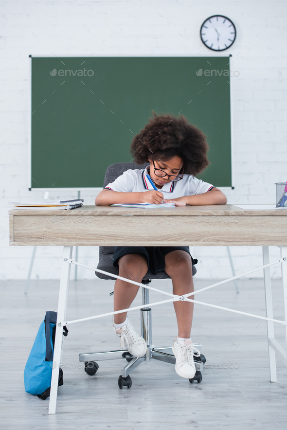 African american pupil in eyeglasses writing on notebook near backpack ...