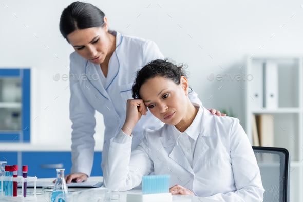 Sad african american scientist sitting near test tubes and colleague on ...