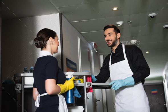 cheerful housekeepers talking near housekeeping cart in corridor of ...