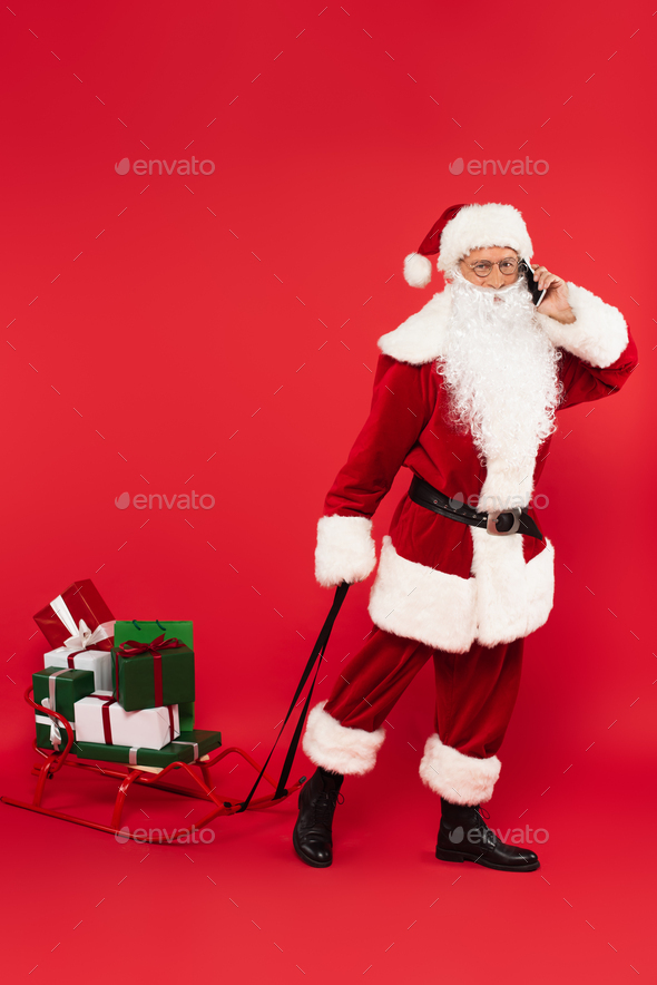 Santa talking on smartphone and pulling sleigh with presents on red ...