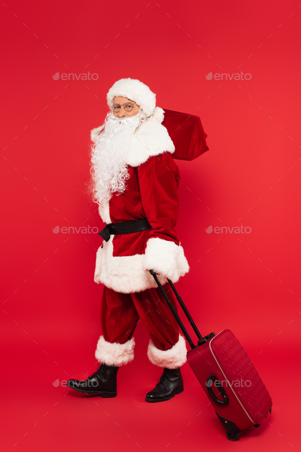 Santa claus in costume holding suitcase and sack on red background ...