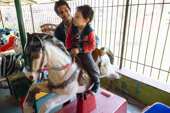 Latino male child riding pretend horse playing with his dad at carousel ...