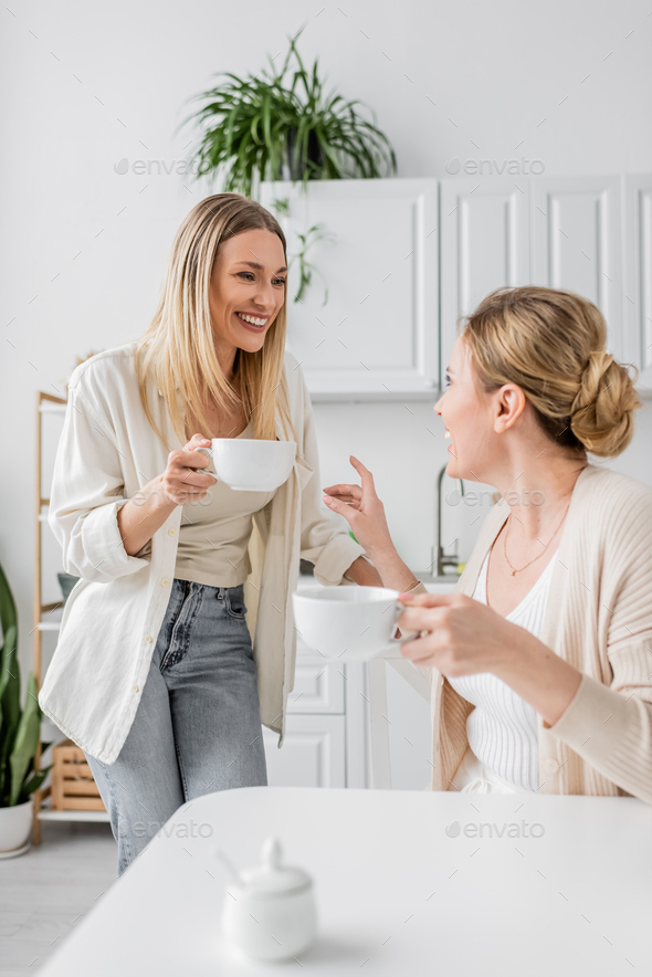 two attractive sisters smiling and looking at each other on kitchen backdrop with plants ...