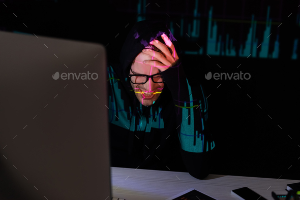 Sad hacker with reflection of charts sitting near devices on black ...