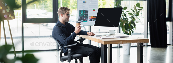 Side view of smiling programmer holding takeaway drink near computer ...
