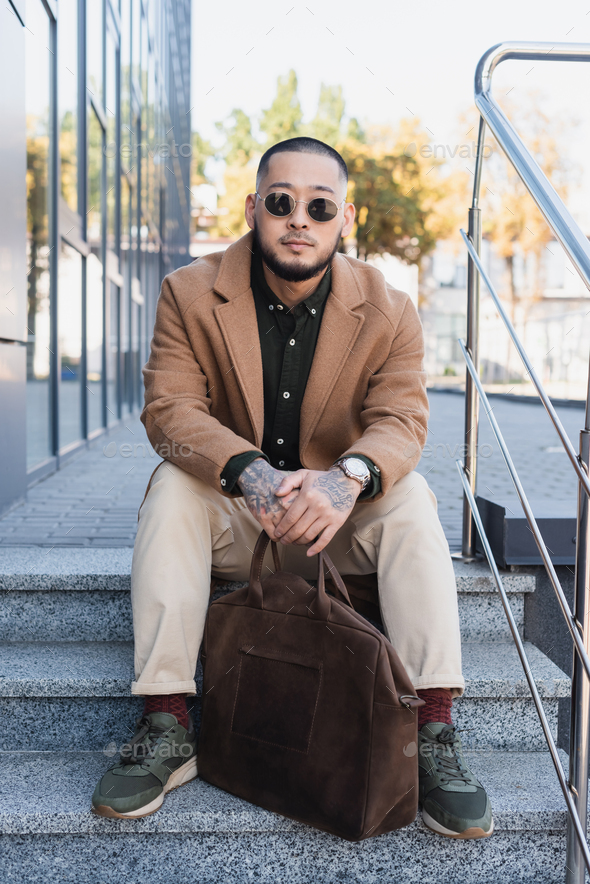 stylish asian man in beige coat and sunglasses sitting on stairs with ...