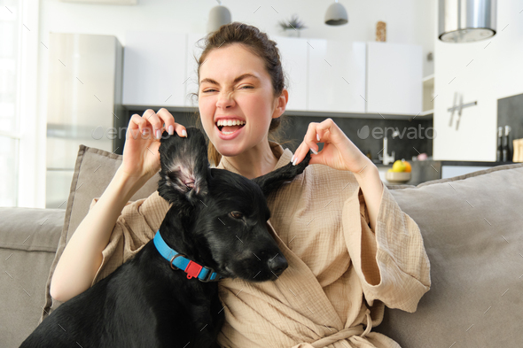 Laughing young woman playing with her dog on sofa. Cute girl smiling ...