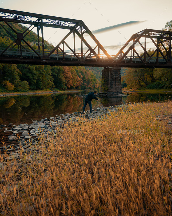 Back view of a man throwing stone in river under a bridge in autumn ...