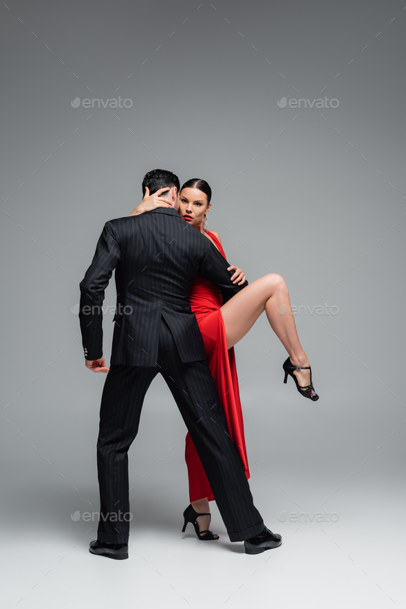 Elegant woman in red dress touching partner while dancing tango on grey ...