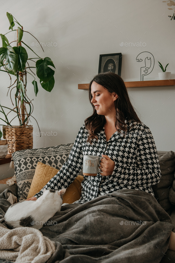 Beautiful woman wearing pajama drinking tea while petting her cat on a ...