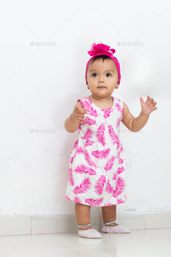 latina baby standing next to a white wall, learning to walk and raising ...