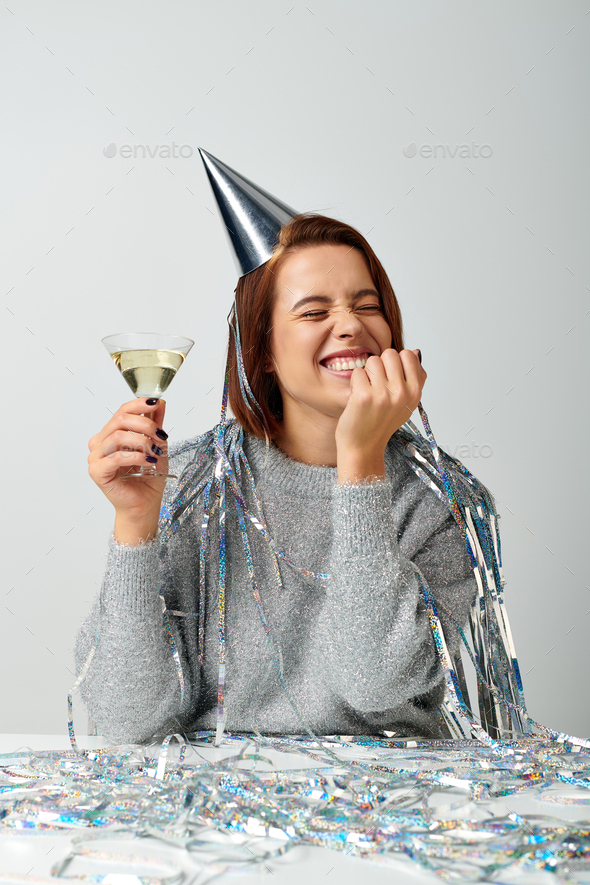 thrilled woman in party cap with tinsel on head holding glass of ...