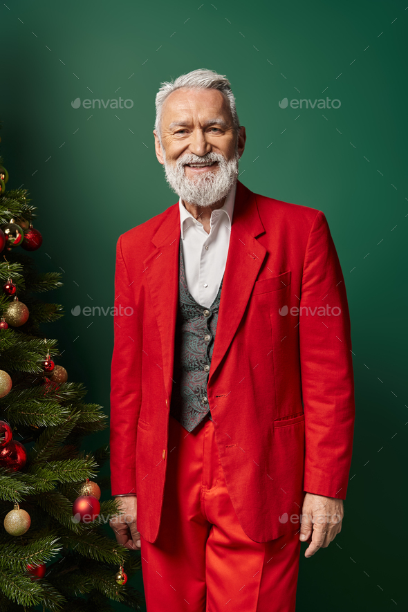 cheerful man in stylish Santa red costume posing near fir tree with ...