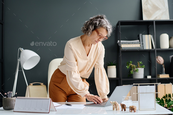 Businesswoman with short curly grey hair bending over desk and laptop ...