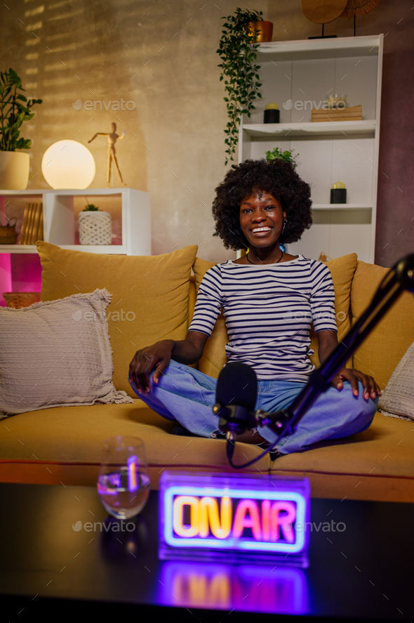 African woman sitting on a yellow sofa in home studio and recording ...