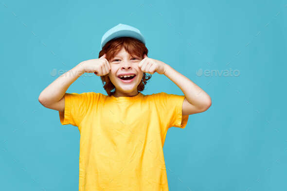 Cheerful boy wide smile cropped view yellow t-shirt cap blue background ...