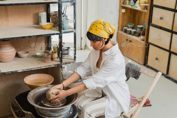 Young asian female artisan in headscarf and workwear molding clay on ...