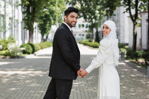happy groom holding hands with smiling muslim bride in wedding dress ...