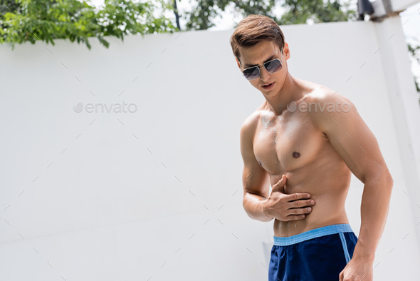 man in sunglasses and swimming trunks applying sunscreen on muscular ...