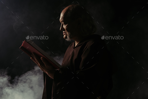 side view of senior priest in cassock reading holy bible on black with ...