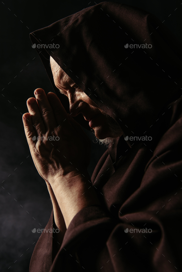 side view of senior priest with praying hands and in black hood on dark ...
