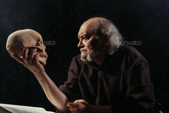senior philosopher in dark robe looking at human skull isolated on ...