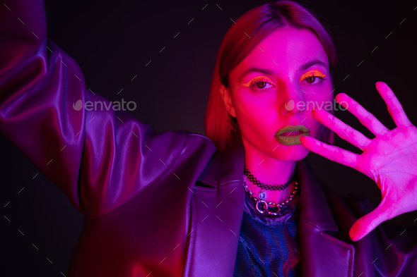 Woman In Leather Jacket And Vibrant Neon Makeup Looking At Camera On Dark Purple Background