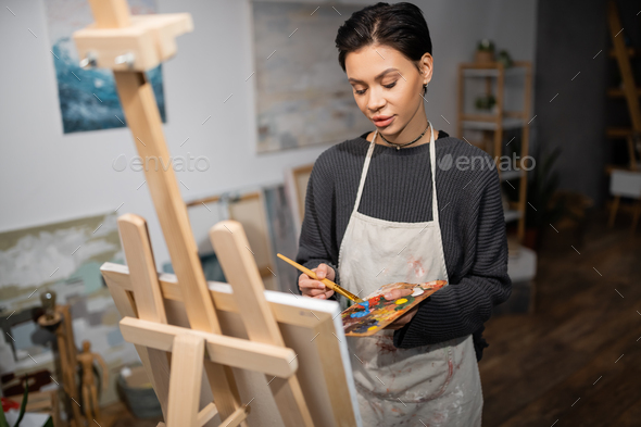 Short haired artist mixing paints on palette near blurred easel in ...