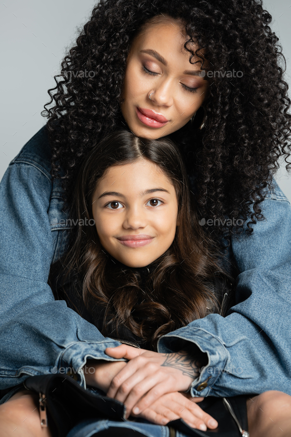 Portrait of curly young mother hugging cheerful child isolated on grey ...