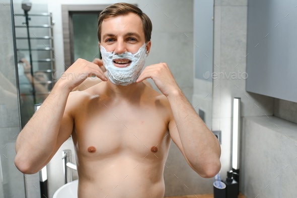 Young smiling Caucasian shirtless man applying shaving foam on face in front of mirror Stock ...