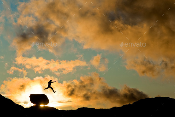Silhouette of a person jumping over a rock on a hill during the sunrise ...