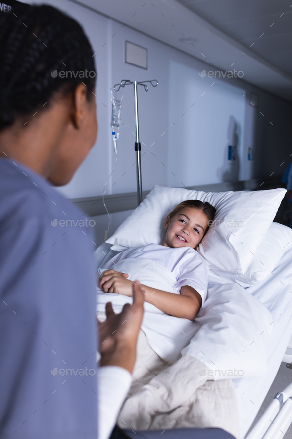 Diverse female doctor talking with happy girl lying in bed in hospital ...