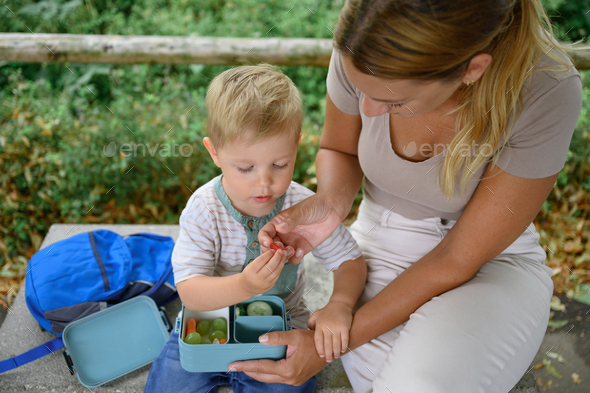 Crop young mother and son sitting on bench with lunchbox in daylight ...